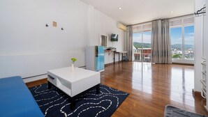 Quarto de hotel moderno com piso de madeira, escrivaninha branca, sofá azul, uma pequena mesa com um vaso, TV na parede e uma grande janela com vista para a cidade.