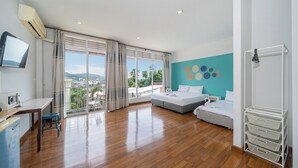 Quarto de hotel moderno com piso de madeira, cama, escrivaninha, cadeira, televisão e varanda com vista para a cidade.