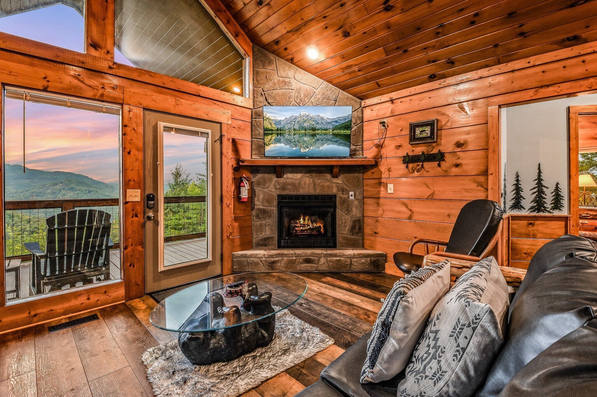Interior acolhedor de uma cabana com lareira de pedra, televisão montada acima e uma grande janela com vista panorâmica.