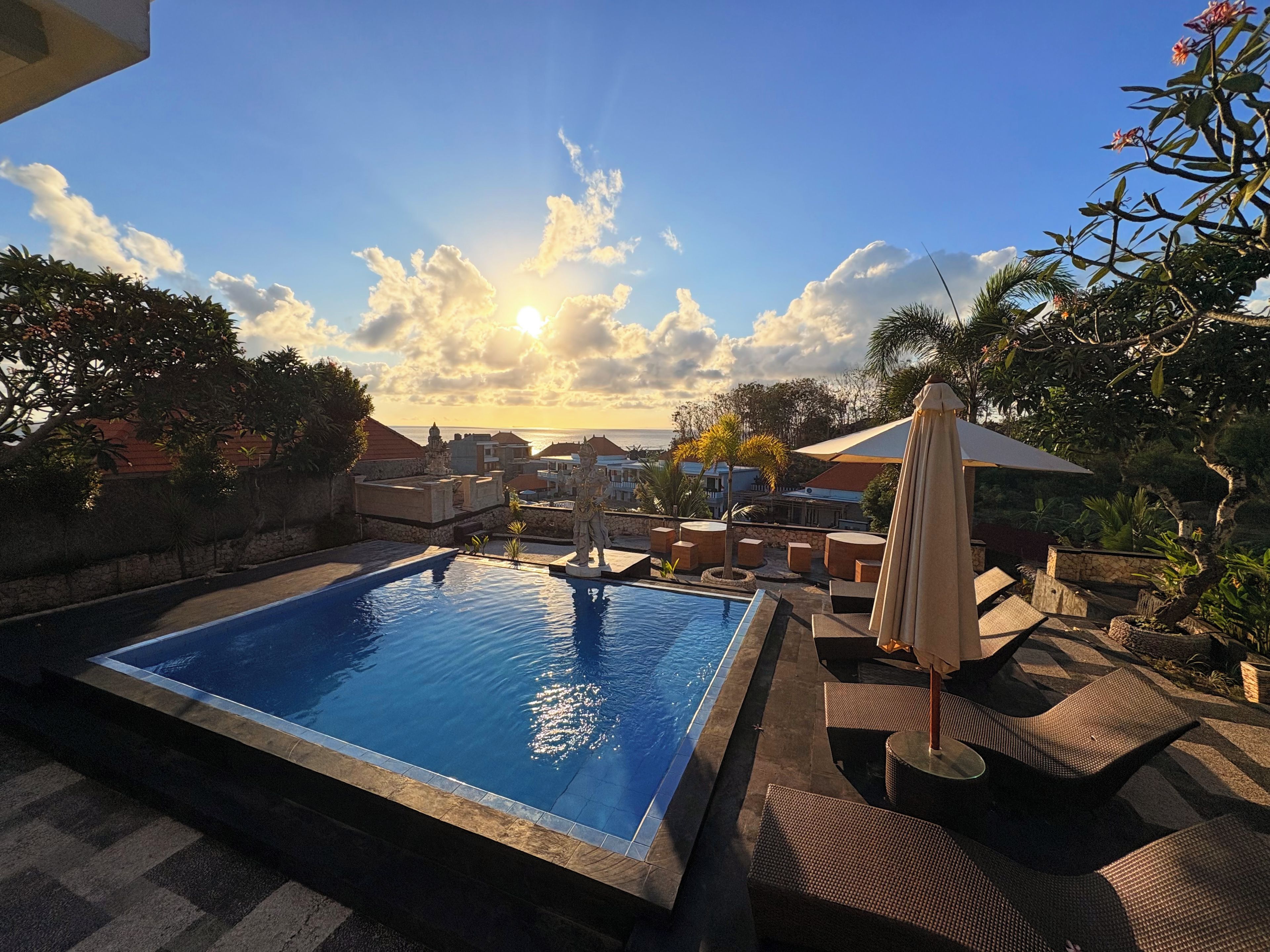 Piscina com espreguiçadeiras e uma estátua, rodeada por vegetação e edifícios.