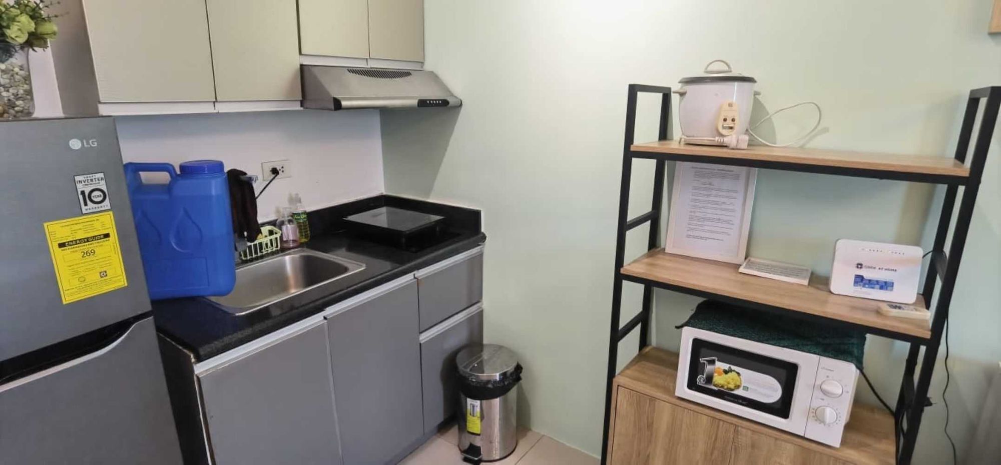 Uma cozinha compacta com geladeira, pia, micro-ondas e uma pequena prateleira com chaleira e relógio digital.