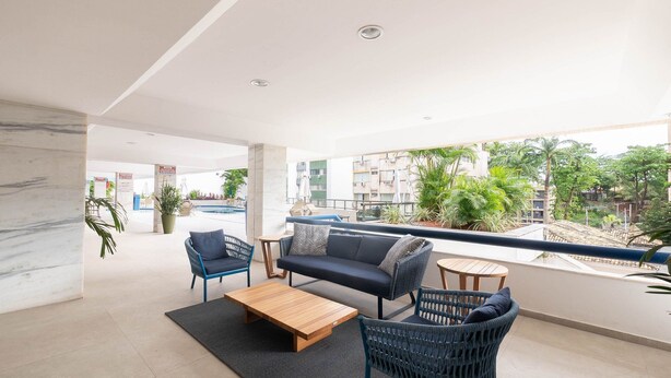 Sala de estar moderna com sofá, duas poltronas e uma mesa de centro. Possui uma varanda com guarda-corpo e vista para a paisagem urbana e áreas verdes.