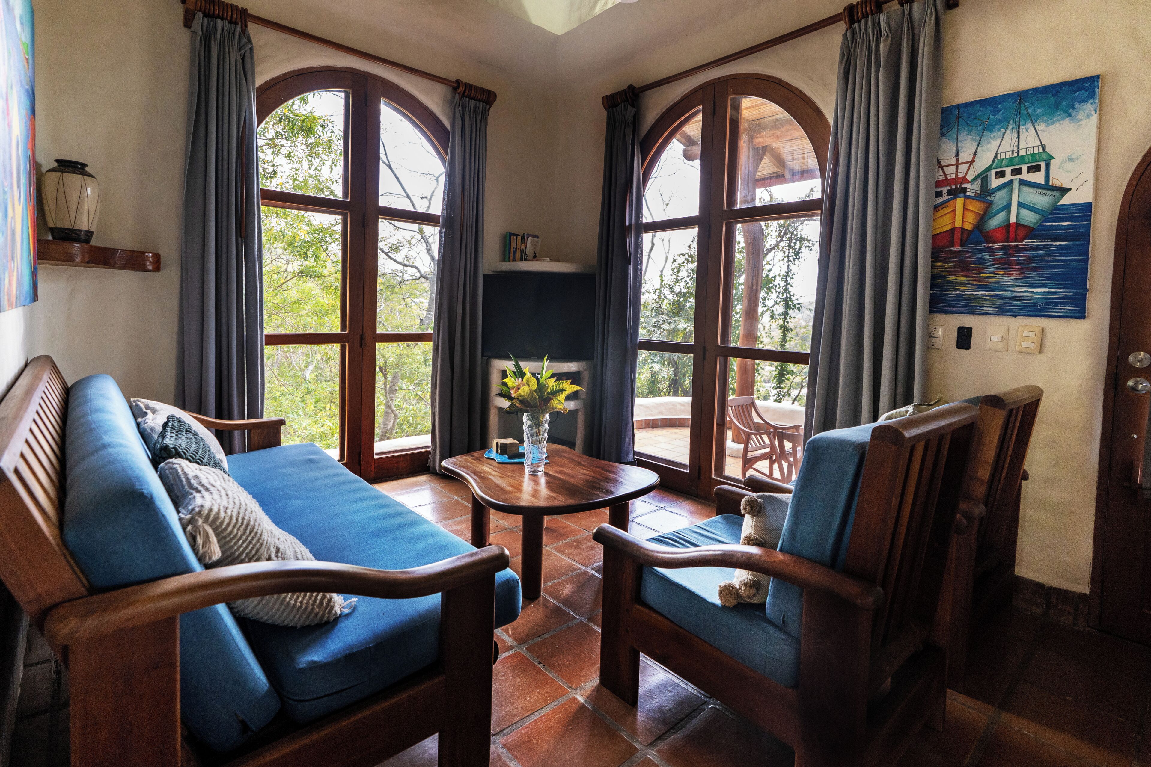 Quarto com sofá azul, cadeiras de madeira, uma pequena mesa com um vaso e janelas amplas com cortinas.