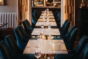 Um restaurante com mesas compridas e cadeiras azul escuras, cada mesa posta com copos de vinho e talheres.