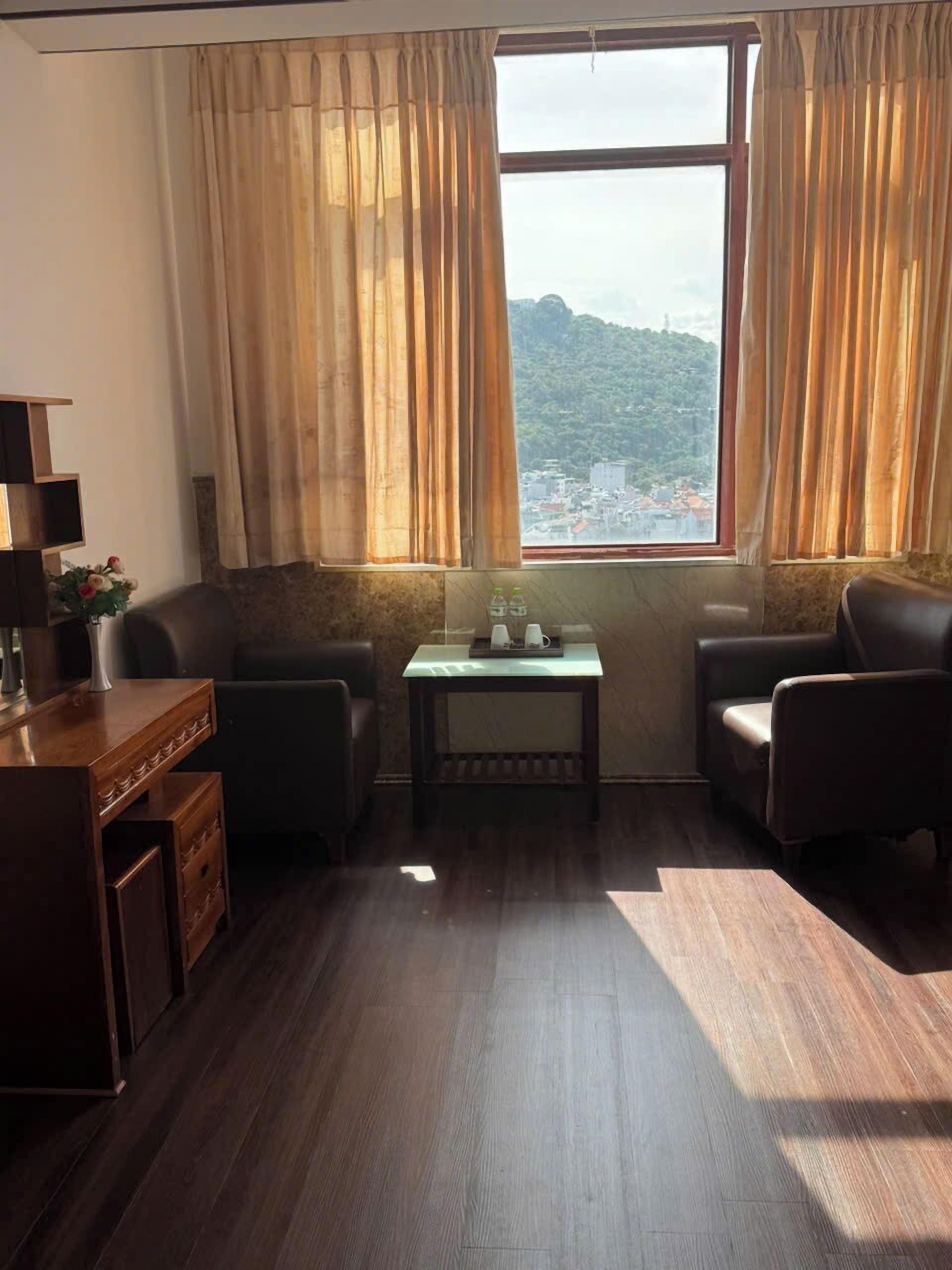 Um quarto com uma escrivaninha de madeira, duas cadeiras, uma mesinha com garrafas e vista para uma paisagem montanhosa através de uma grande janela com cortinas.