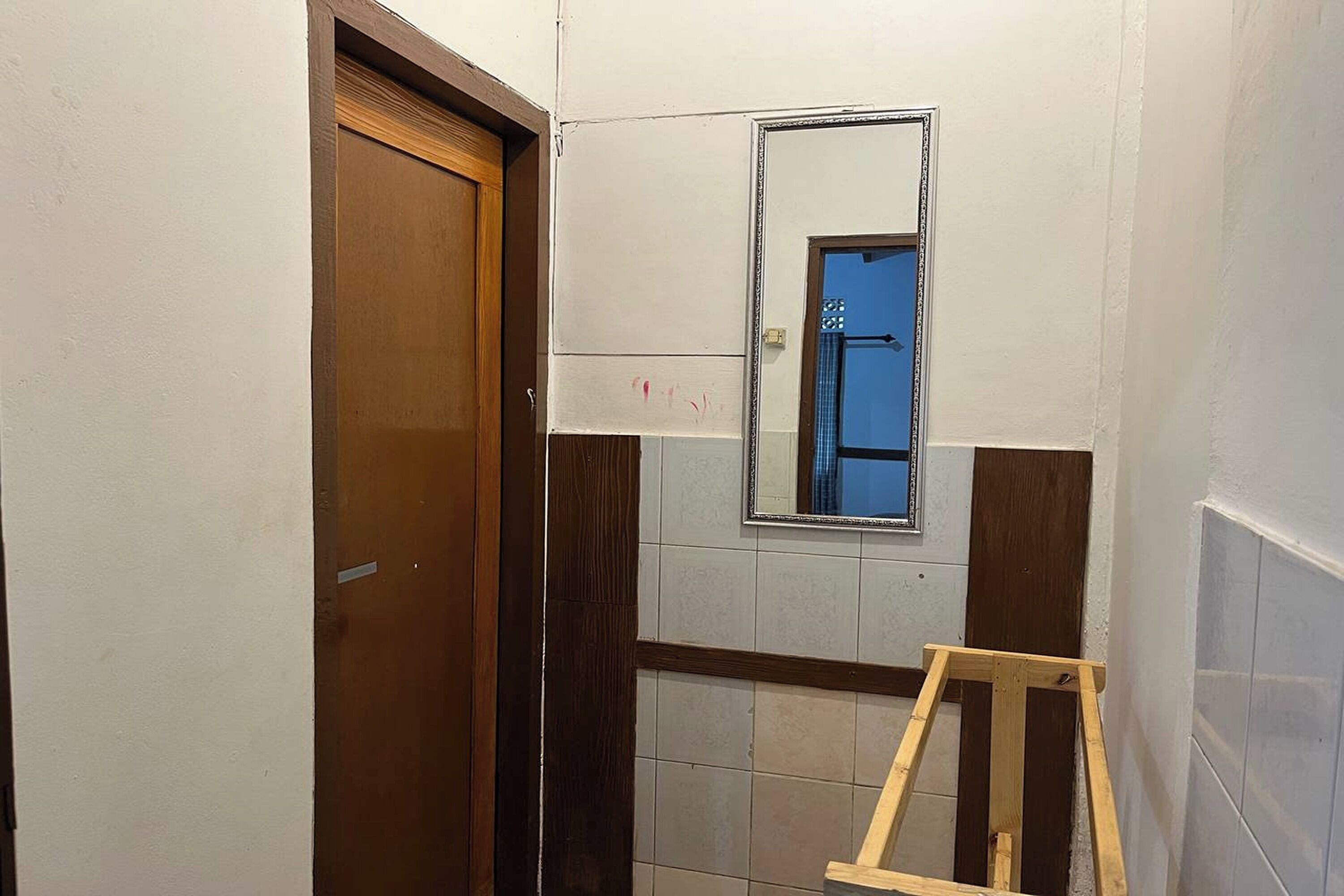 Um corredor estreito com porta de madeira, uma pequena janela com espelho e uma escada de madeira encostada à parede.