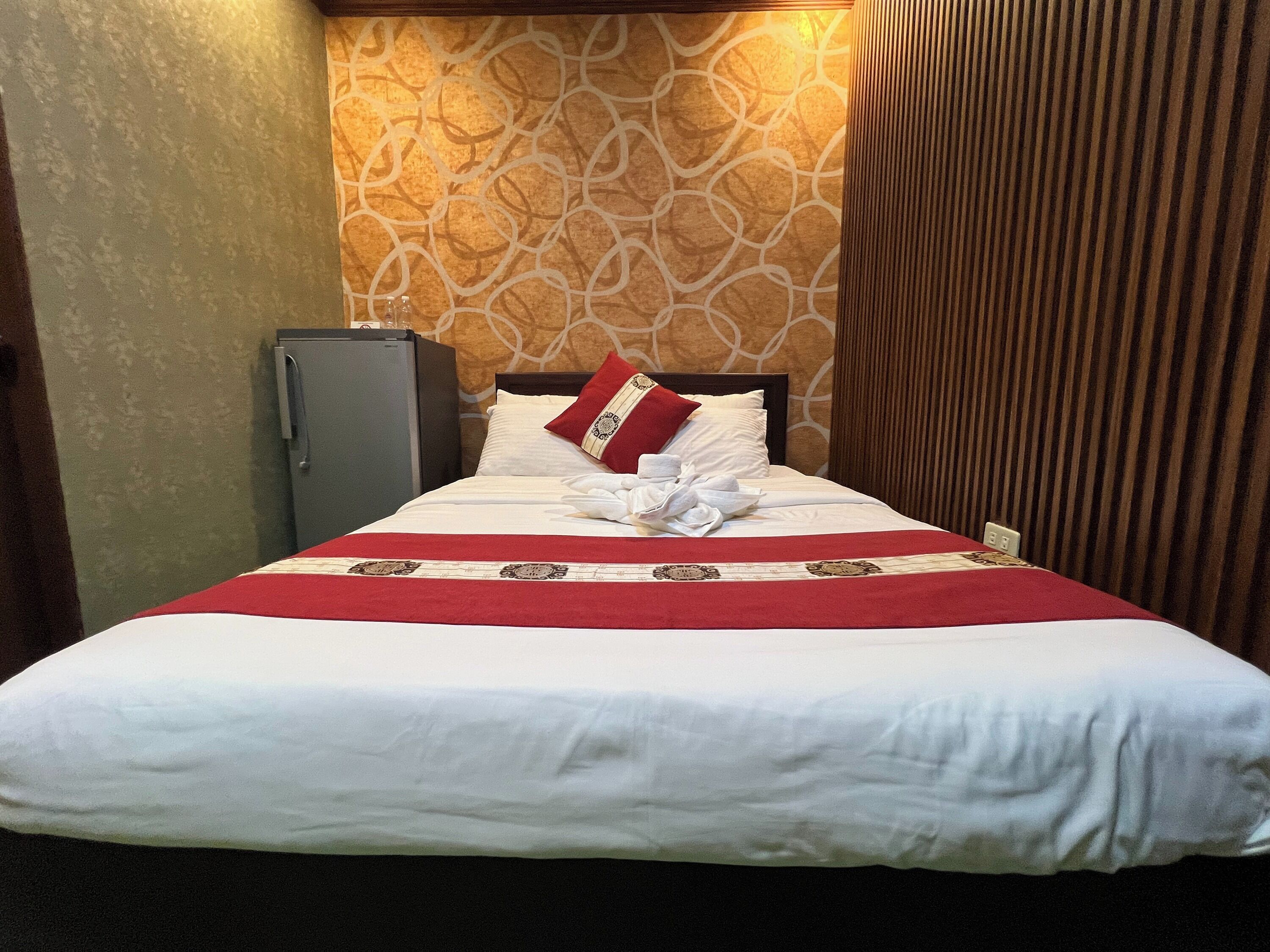 Uma cama bem arrumada com lençóis brancos e uma almofada vermelha decorativa, um pequeno frigorífico à esquerda e um papel de parede estampado atrás da cama.