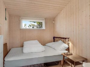 Quarto pequeno com cama de solteiro, teto e paredes de madeira, janela com vista para árvores, mesa de cabeceira com um livro e tomada na parede.