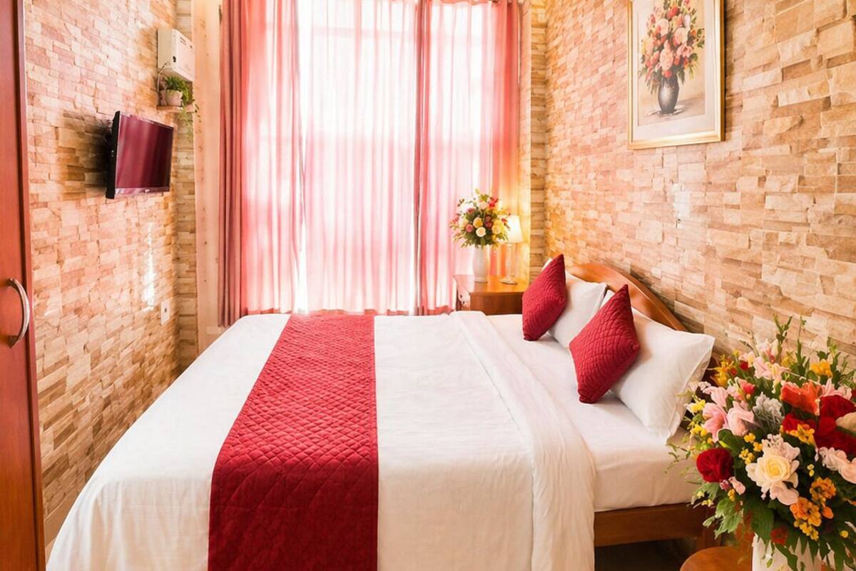 Quarto com cama, ventoinha no teto, janela com cortinas, televisão fixada na parede, mesa de cabeceira com abajur e um vaso de flores.
