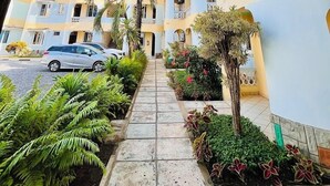 Um edifício de vários andares com varandas, um caminho pavimentado e vegetação exuberante, incluindo palmeiras e samambaias.