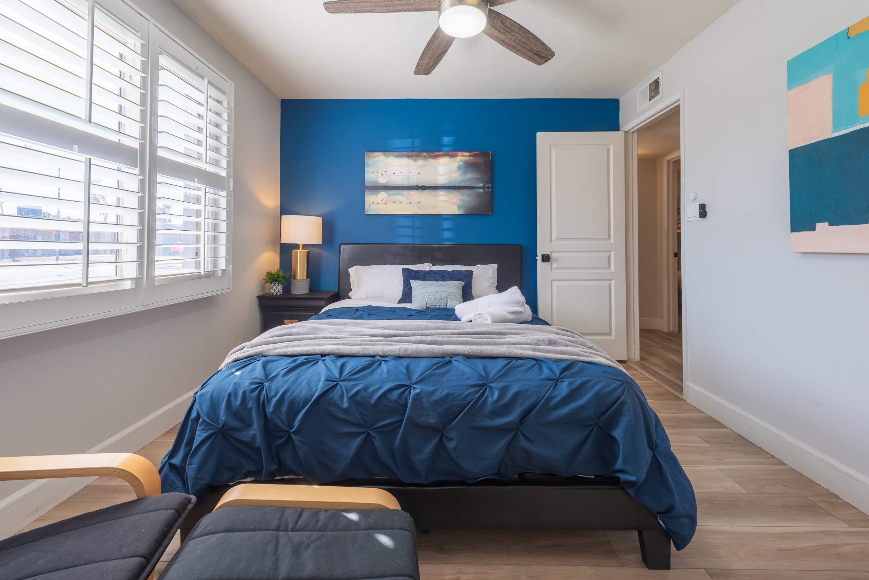 Quarto com cama, ventilador de teto, mesa de cabeceira, abajur e janela com persianas.