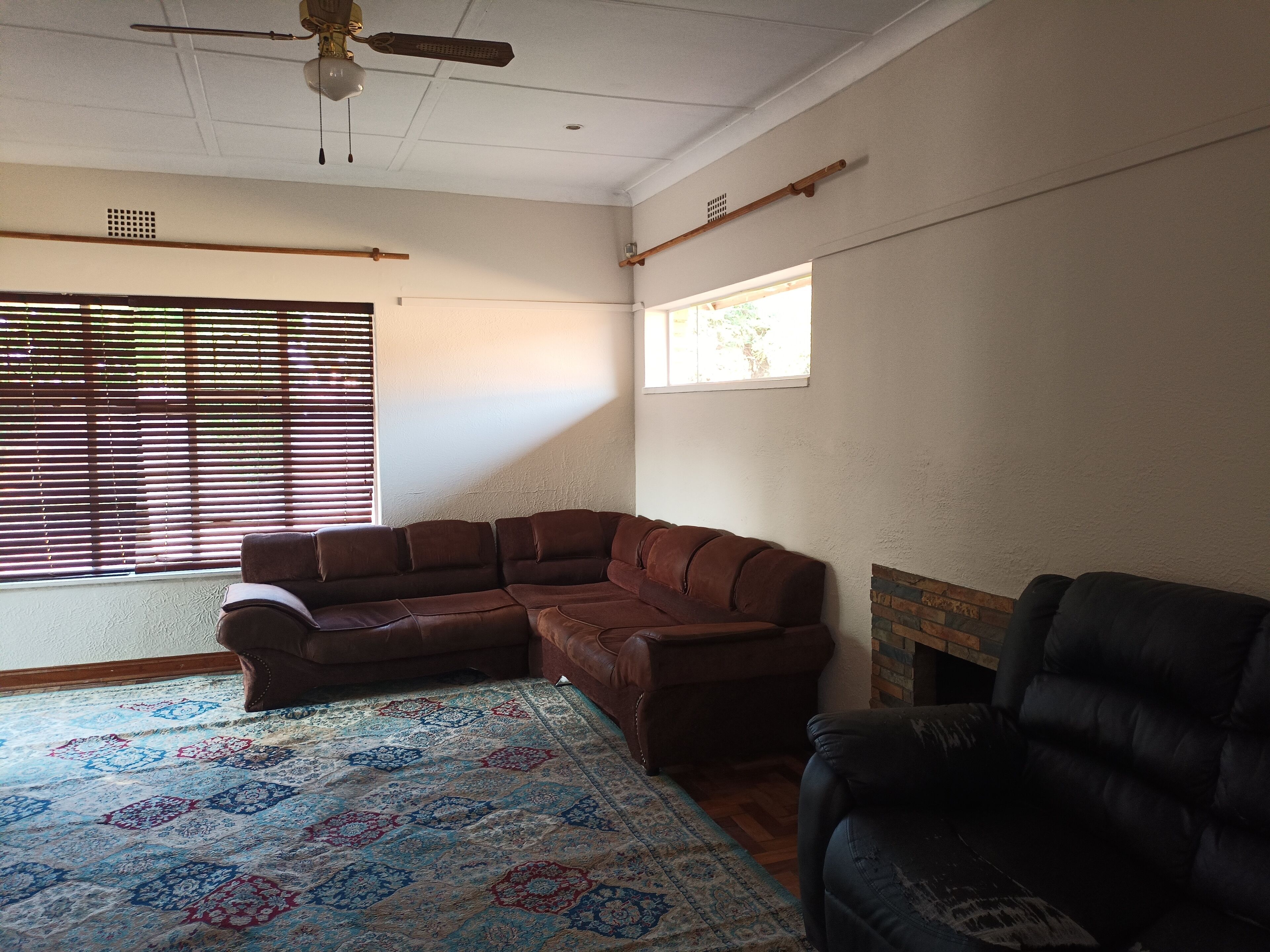 Uma sala de estar com sofá seccional, uma poltrona, tapete estampado, ventilador de teto e janela com persianas.