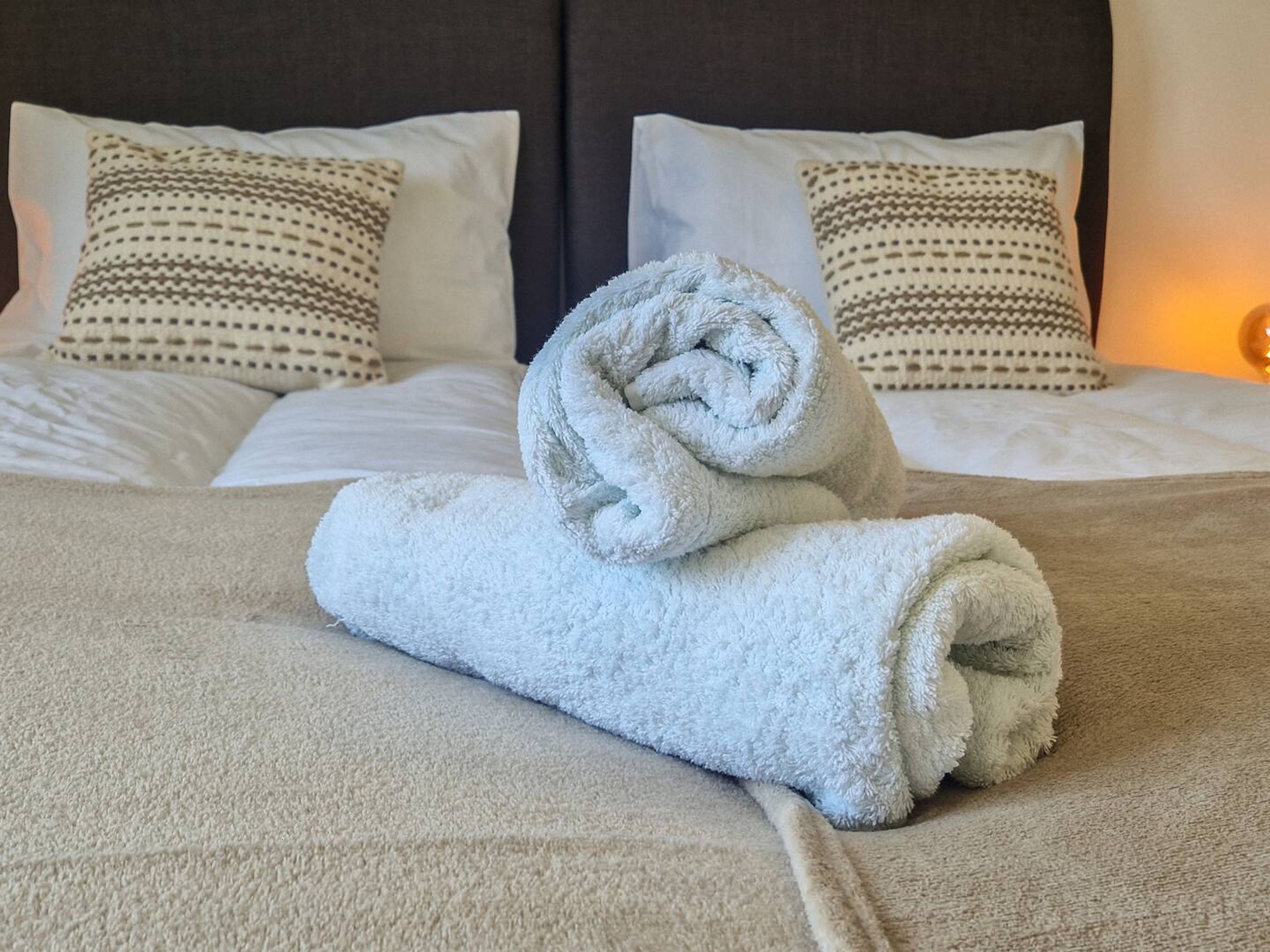 Cama bem arrumada com uma toalha branca enrolada e almofadas estampadas.