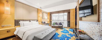 Quarto de hotel com cama, sofá, televisão, mesa pequena e janela com cortinas.
