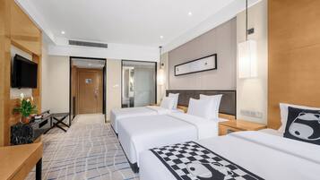 Quarto de hotel com uma cama grande, uma cama menor, uma escrivaninha, uma televisão e um banheiro visível através de uma porta de vidro.