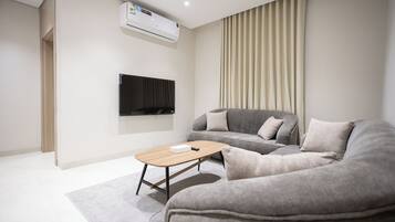 Uma sala de estar moderna com um sofá cinza, uma mesa de centro de madeira, uma TV na parede e uma janela grande com cortinas.