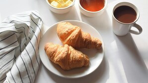 Um prato com dois croissants, uma tigela de cubos de manteiga e duas chávenas de chá sobre uma mesa branca.