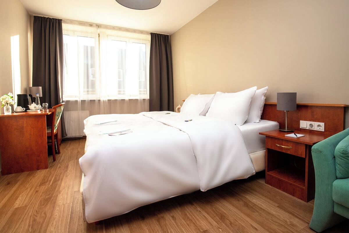 Quarto de hotel com cama, escrivaninha, cadeira, abajur e janela com cortinas.