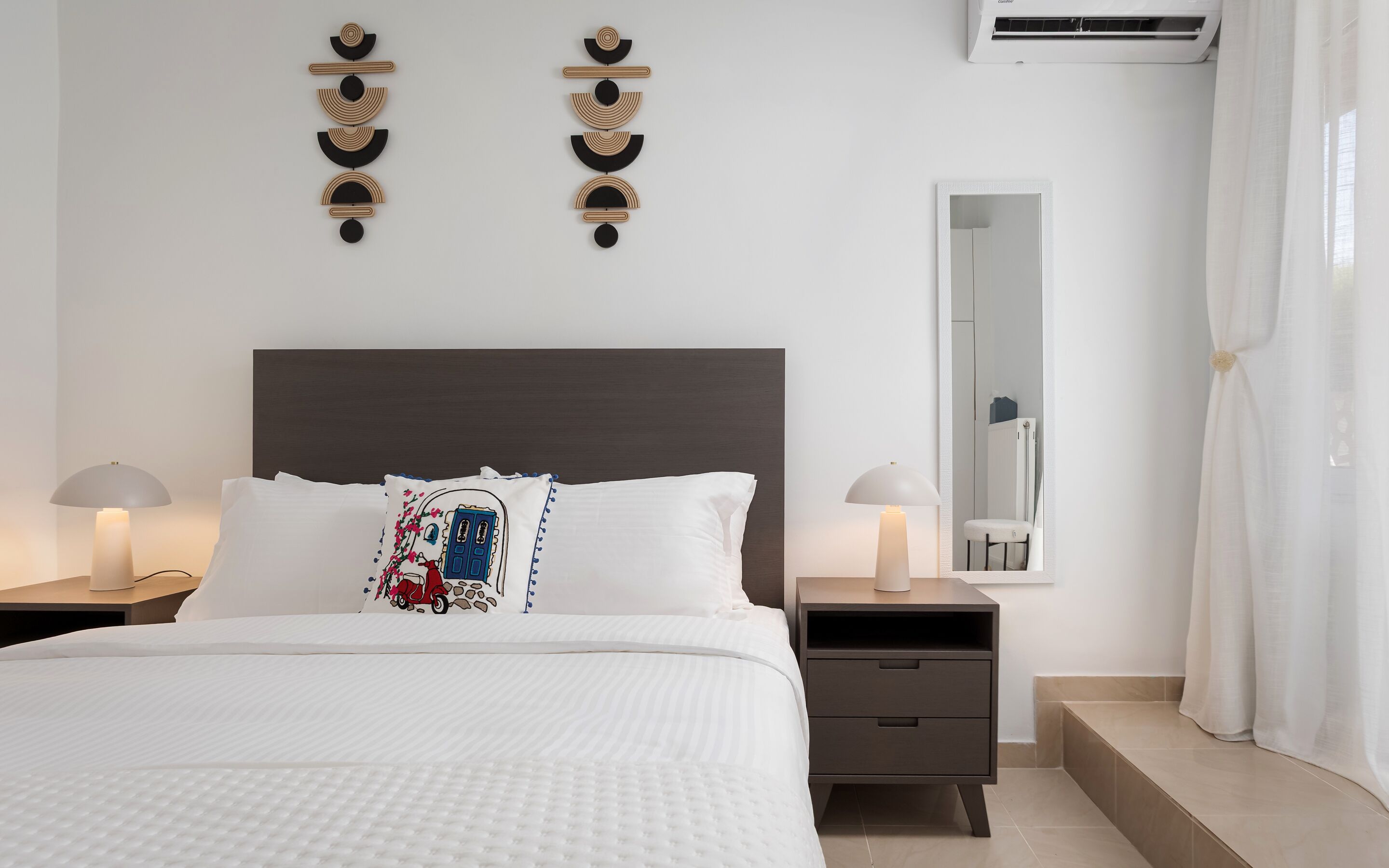 Quarto de hotel moderno, com uma cama grande, duas mesas de cabeceira com abajures, um espelho fixado na parede e um detalhe decorativo na parede.