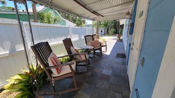 Um alpendre coberto com cadeiras de balanço de madeira e uma pequena mesa, cercado por um muro branco e plantas em vasos.