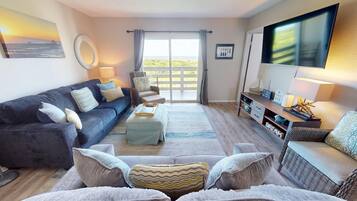 Sala de estar com um sofá azul escuro, um banquinho azul claro e um sofá cinza. Há uma TV na parede, uma janela com cortinas e vista para o exterior.