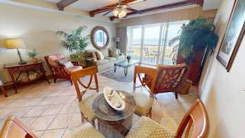 Uma sala de estar ampla com área de jantar, uma janela grande com vista para o oceano e vários elementos de decoração tropical.