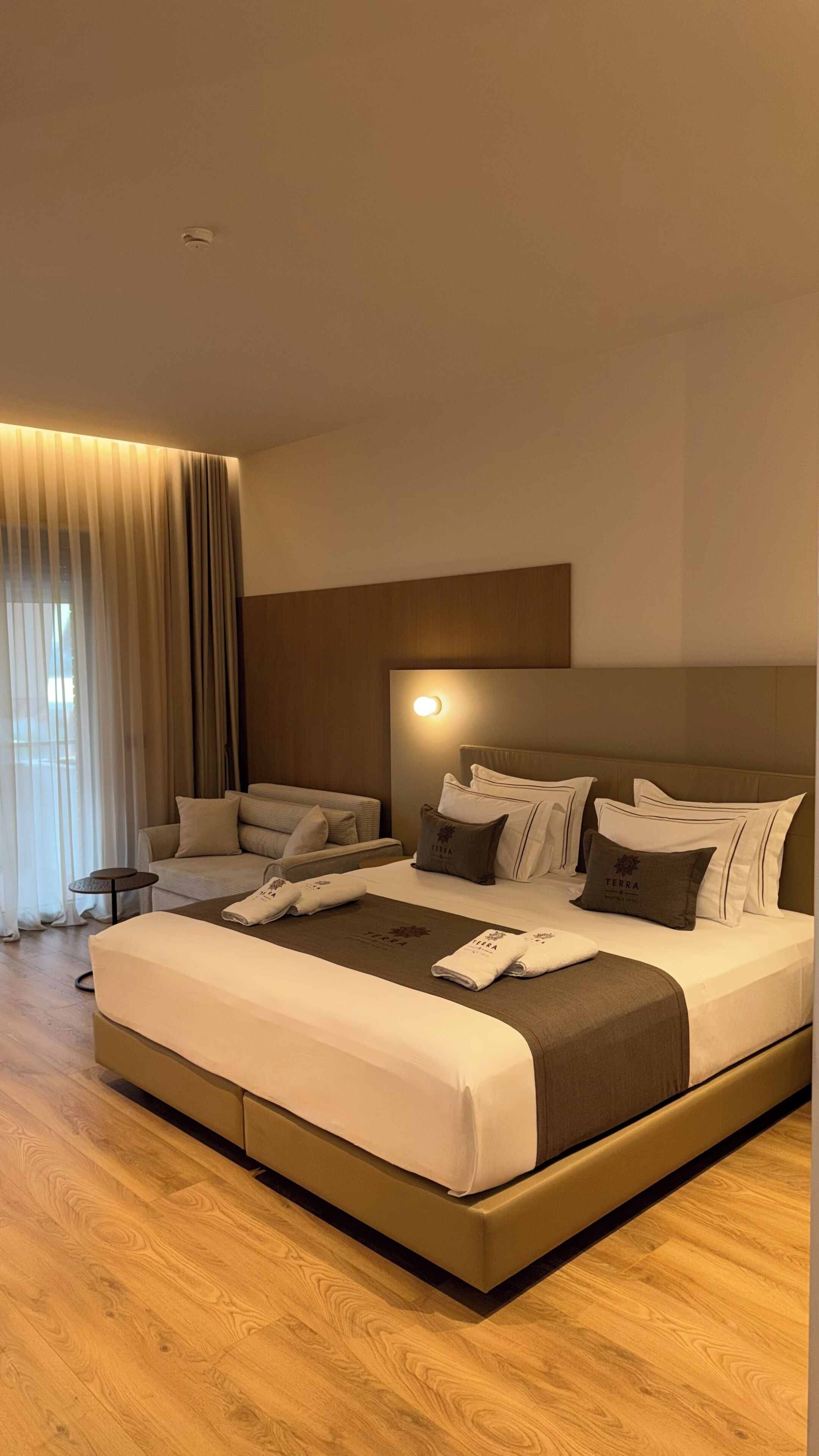 Quarto de hotel moderno com cama grande, mesa de cabeceira, sofá e janela com cortinas.