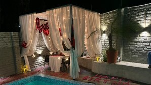 Uma área de piscina com toldo branco e cortinas esvoaçantes, uma pequena mesa e uma cadeira.