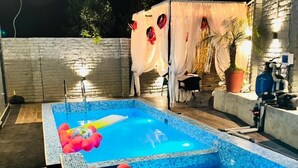 Piscina retangular com borda azulejada, rodeada por um muro de pedra e uma área de estar coberta com cortinas brancas.