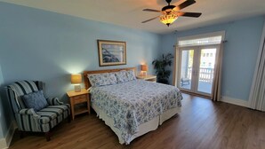 Quarto com cama, cadeira, mesa de cabeceira, ventilador de teto e porta que dá para uma varanda.