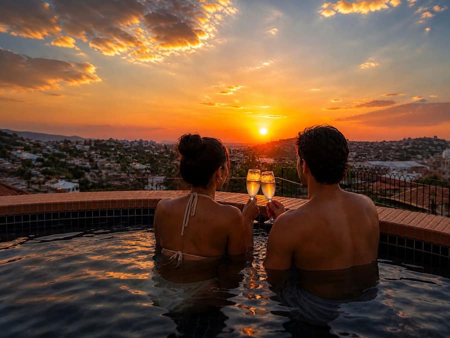 Um casal em banheira de hidromassagem com vista para o horizonte urbano ao pôr do sol.