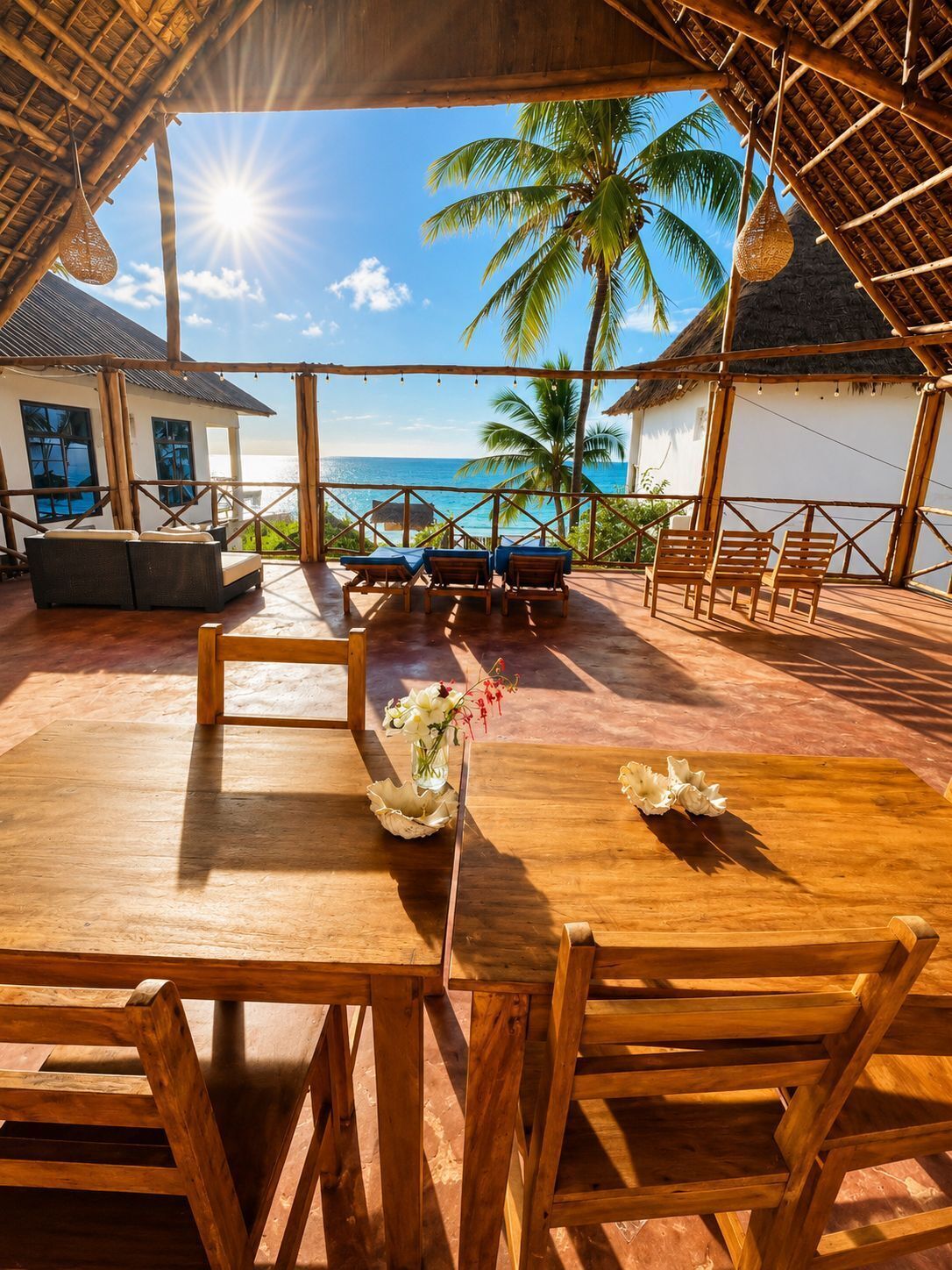Um deck de madeira com uma mesa e cadeiras, com vista para uma praia com palmeiras e o oceano.