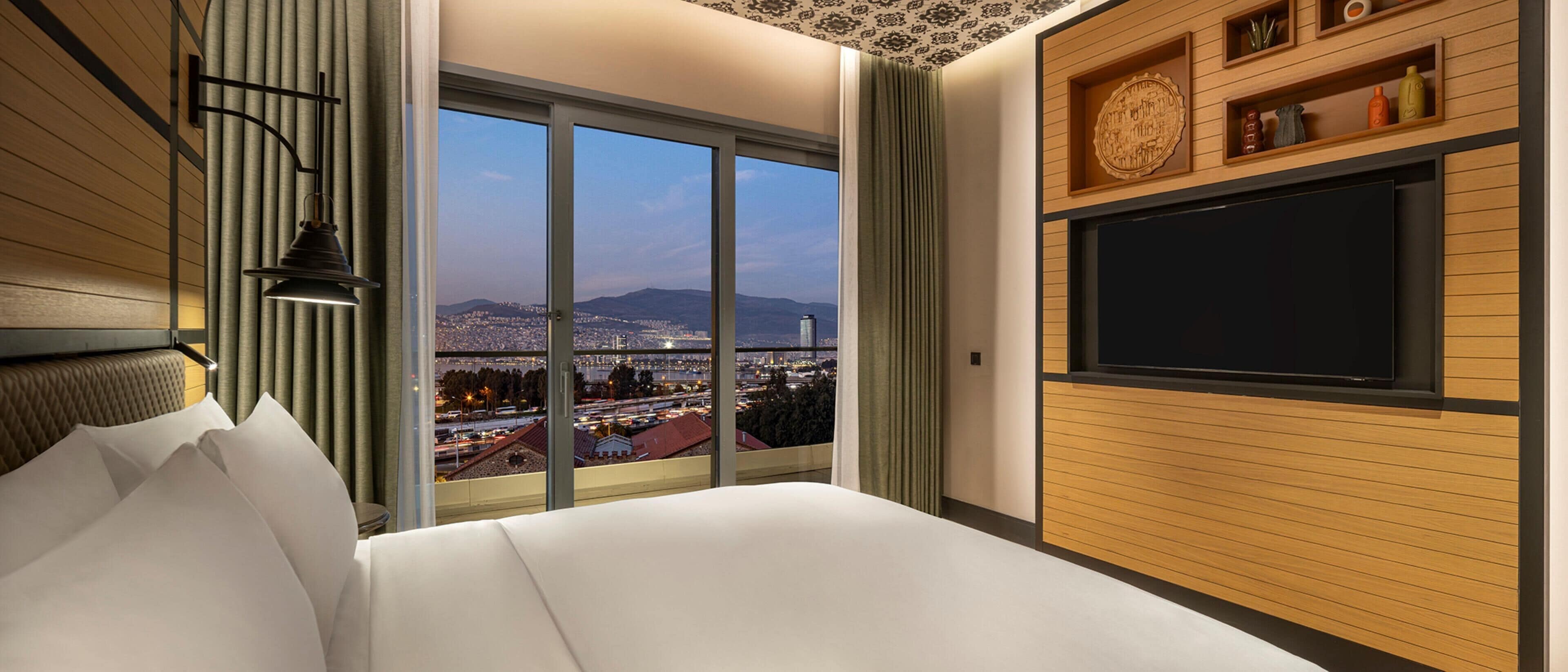 Quarto de hotel com uma cama grande, vista para a cidade através de janelas do chão ao teto, televisão de tela plana na parede e painéis de madeira nas paredes.