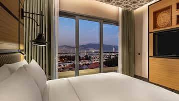 Quarto de hotel com uma cama grande, vista para a cidade através de janelas do chão ao teto, televisão de tela plana na parede e painéis de madeira nas paredes.