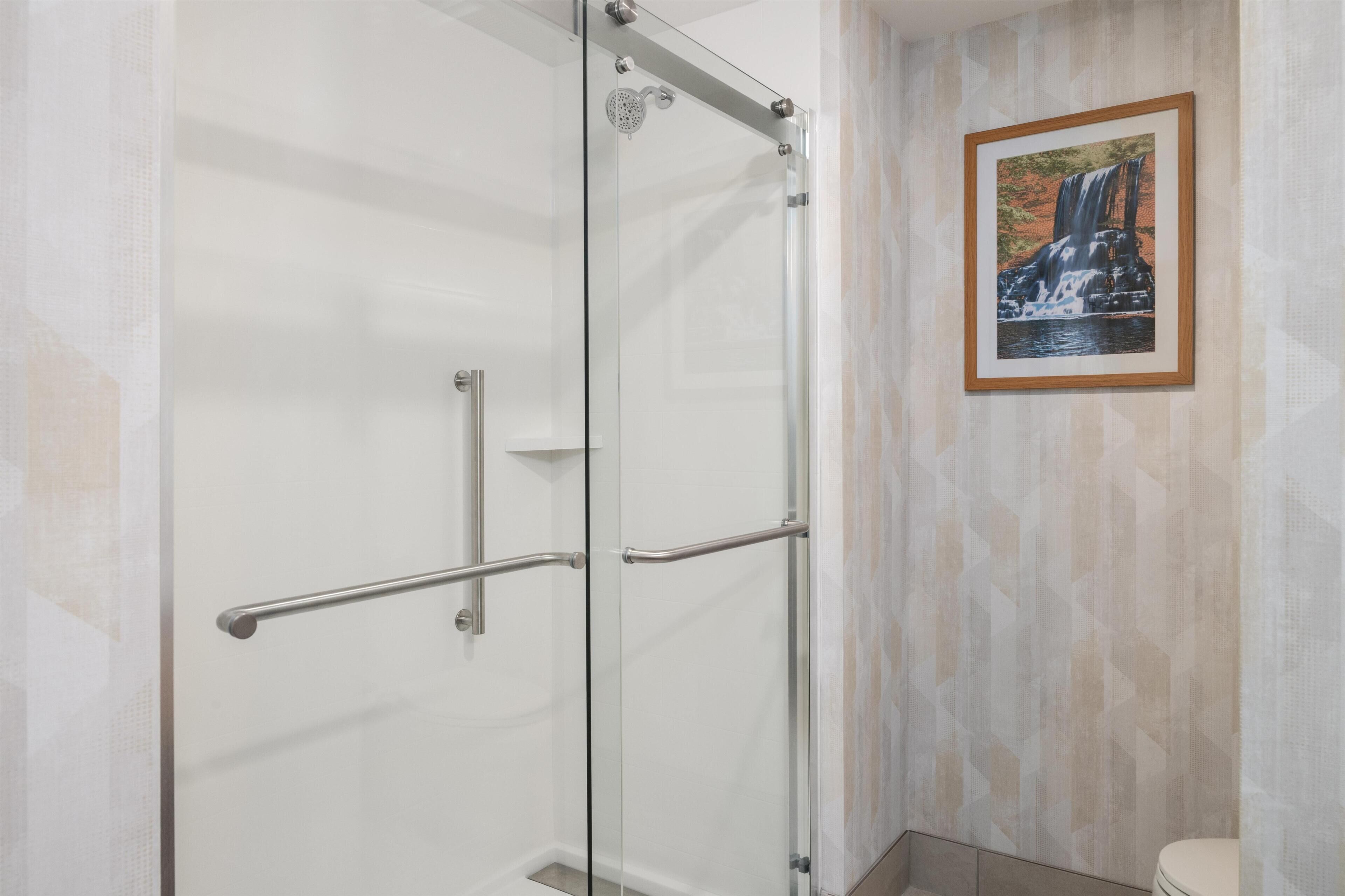 Chuveiro envidraçado com barras de apoio, quadro de uma cascata na parede e um vaso sanitário.
