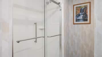 Chuveiro envidraçado com barras de apoio, quadro de uma cascata na parede e um vaso sanitário.