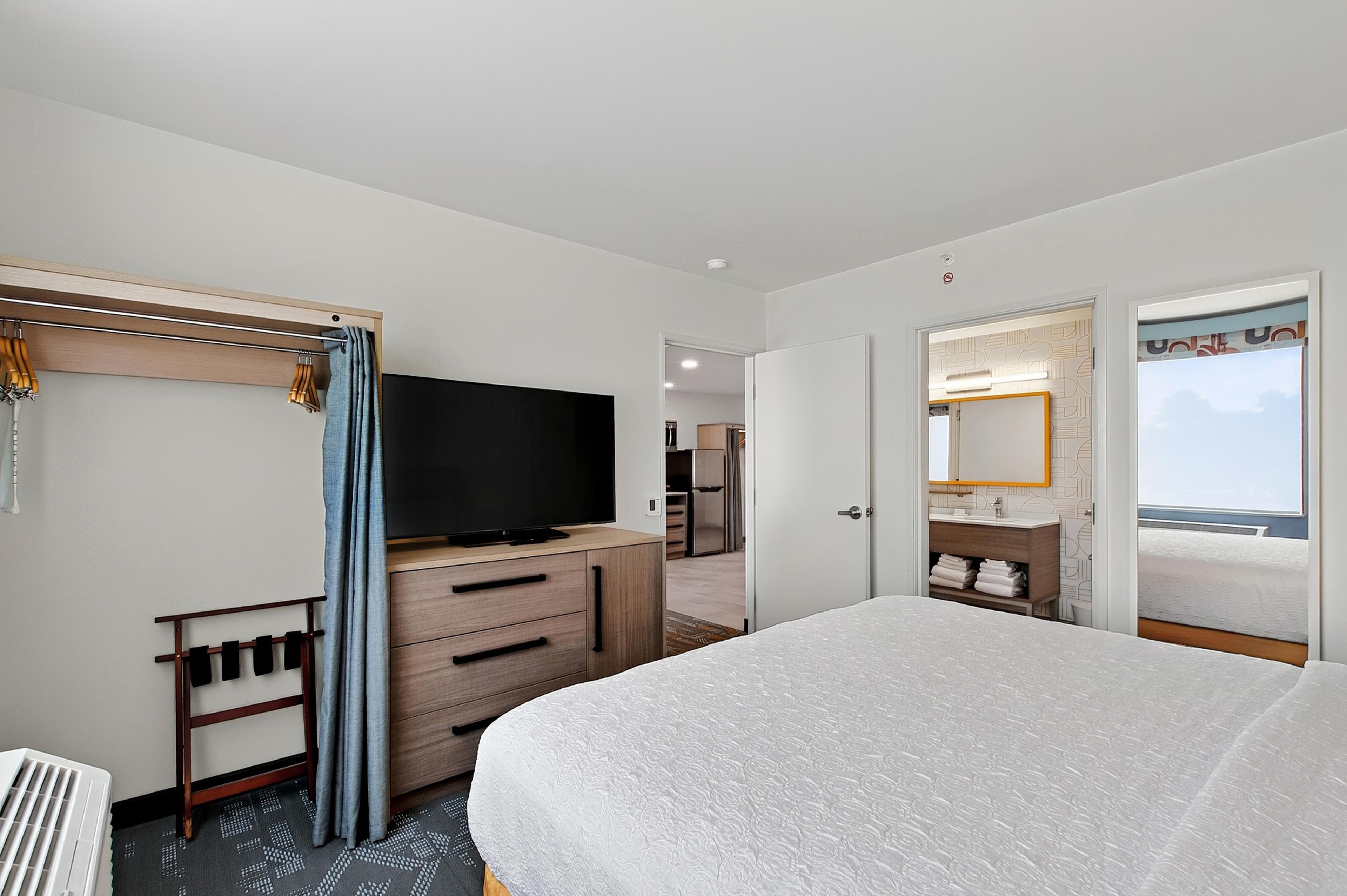 Quarto de hotel com cama, televisão, guarda-roupa e banheiro visível através de uma porta aberta.