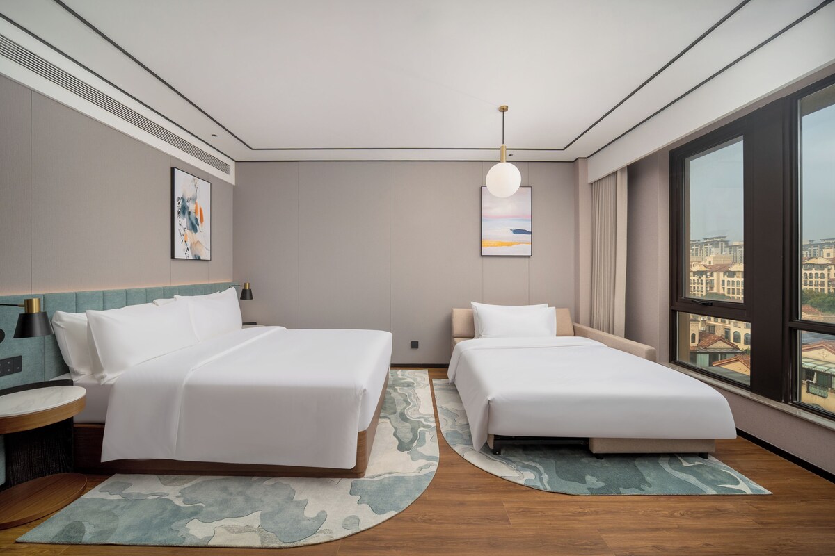Quarto de hotel com duas camas, uma mesa redonda, uma janela grande com vista para a cidade e obras de arte nas paredes.