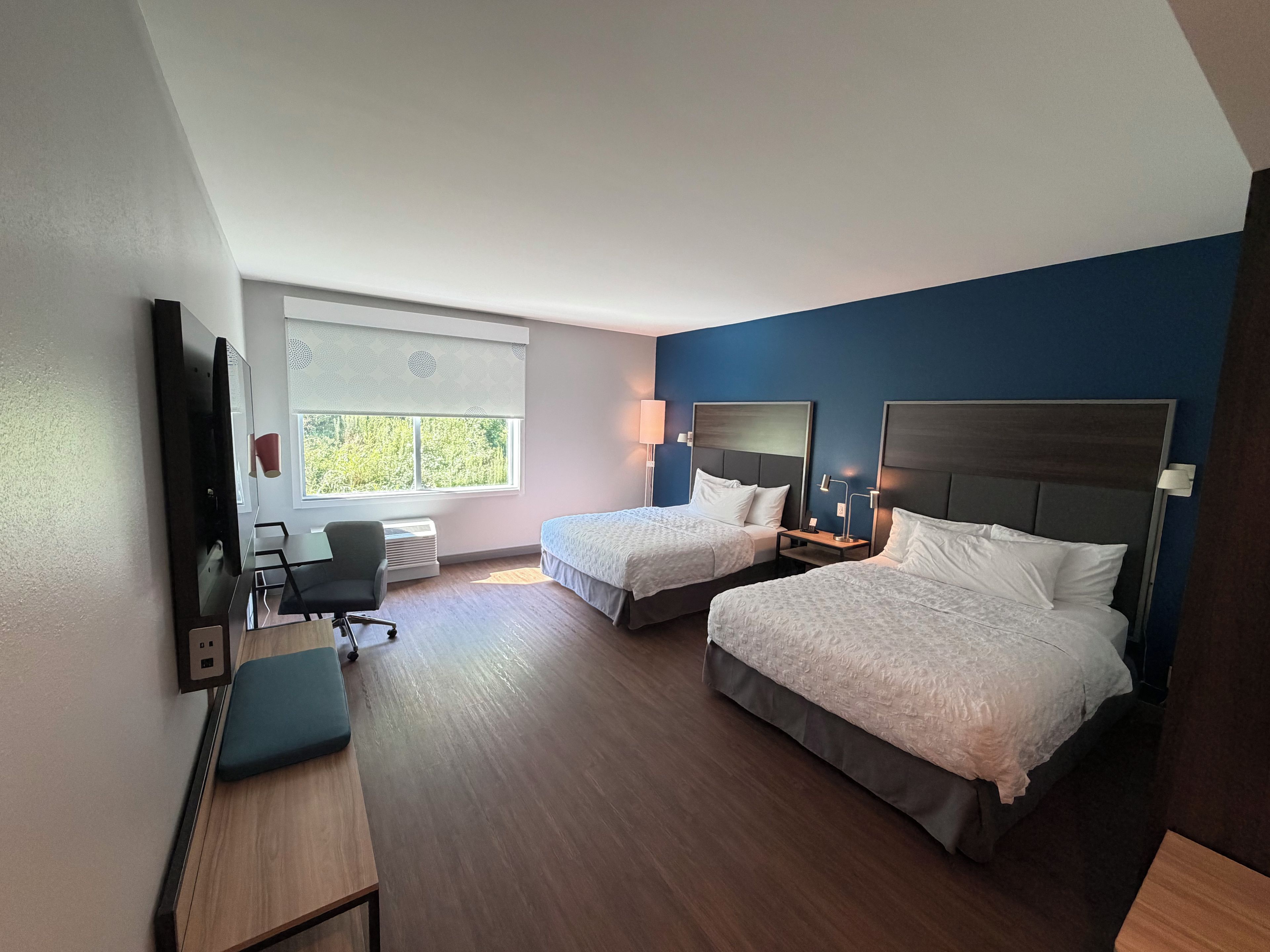 Quarto de hotel com duas camas, uma mesa, uma cadeira, uma televisão e uma janela com persianas.