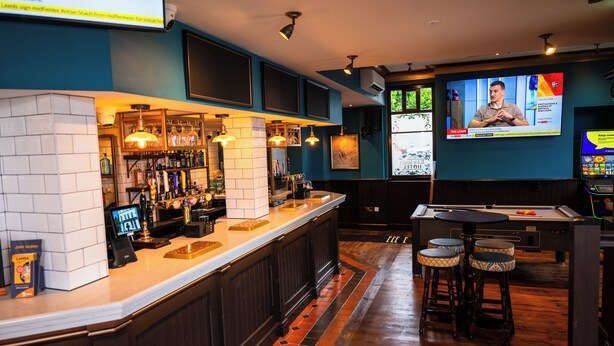 Uma área de bar com balcão extenso, banquetas e uma tela de televisão exibindo um programa de notícias.