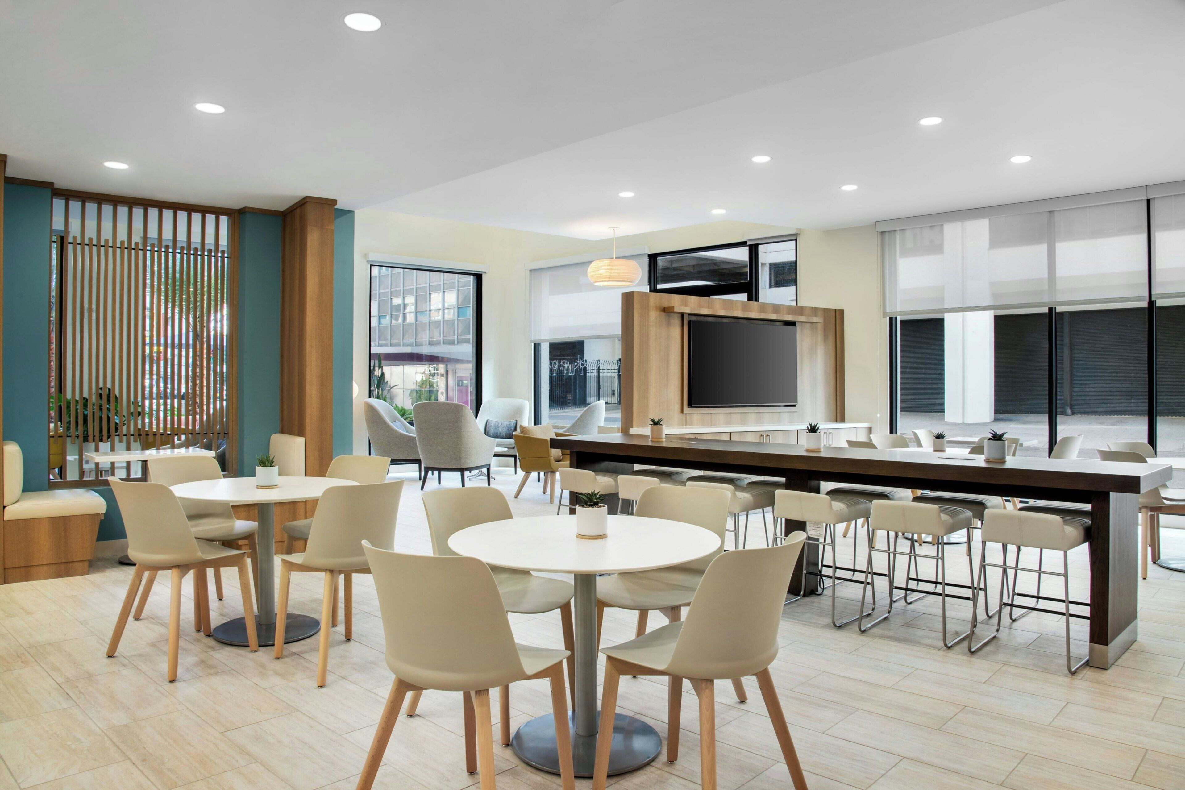 Um hall moderno de hotel com mesas redondas, banquetas de bar e uma grande tela de televisão.