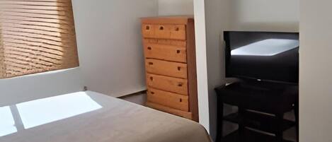 Quarto com cama, cômoda de madeira, televisão e janela com persianas.