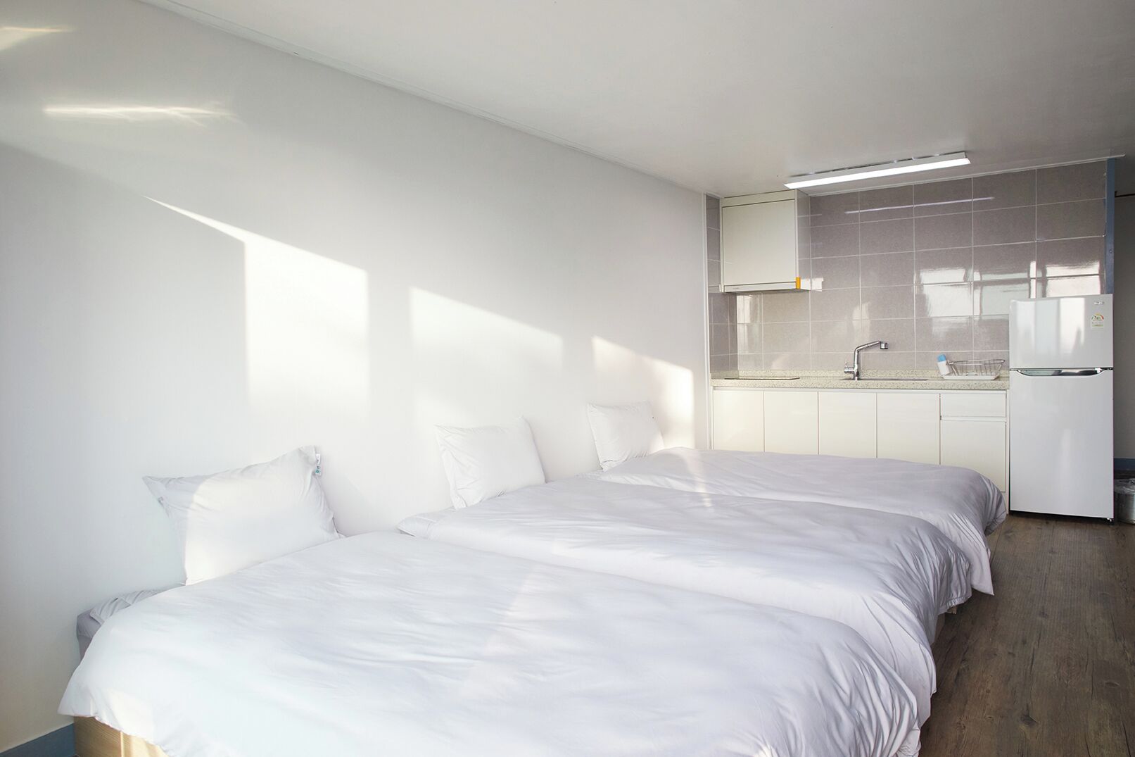 Quarto moderno e minimalista com uma cama grande, uma kitchenette pequena e um ambiente claro e iluminado pelo sol.
