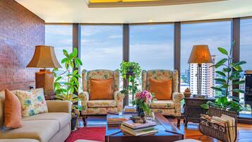 Uma sala de estar espaçosa com dois poltronas florais, um sofá, uma mesa de centro com livros e um vaso, e grandes janelas com vista para a cidade.