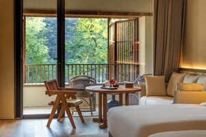 Quarto de hotel com cama, sofá, mesa de jantar e varanda com vista para árvores.