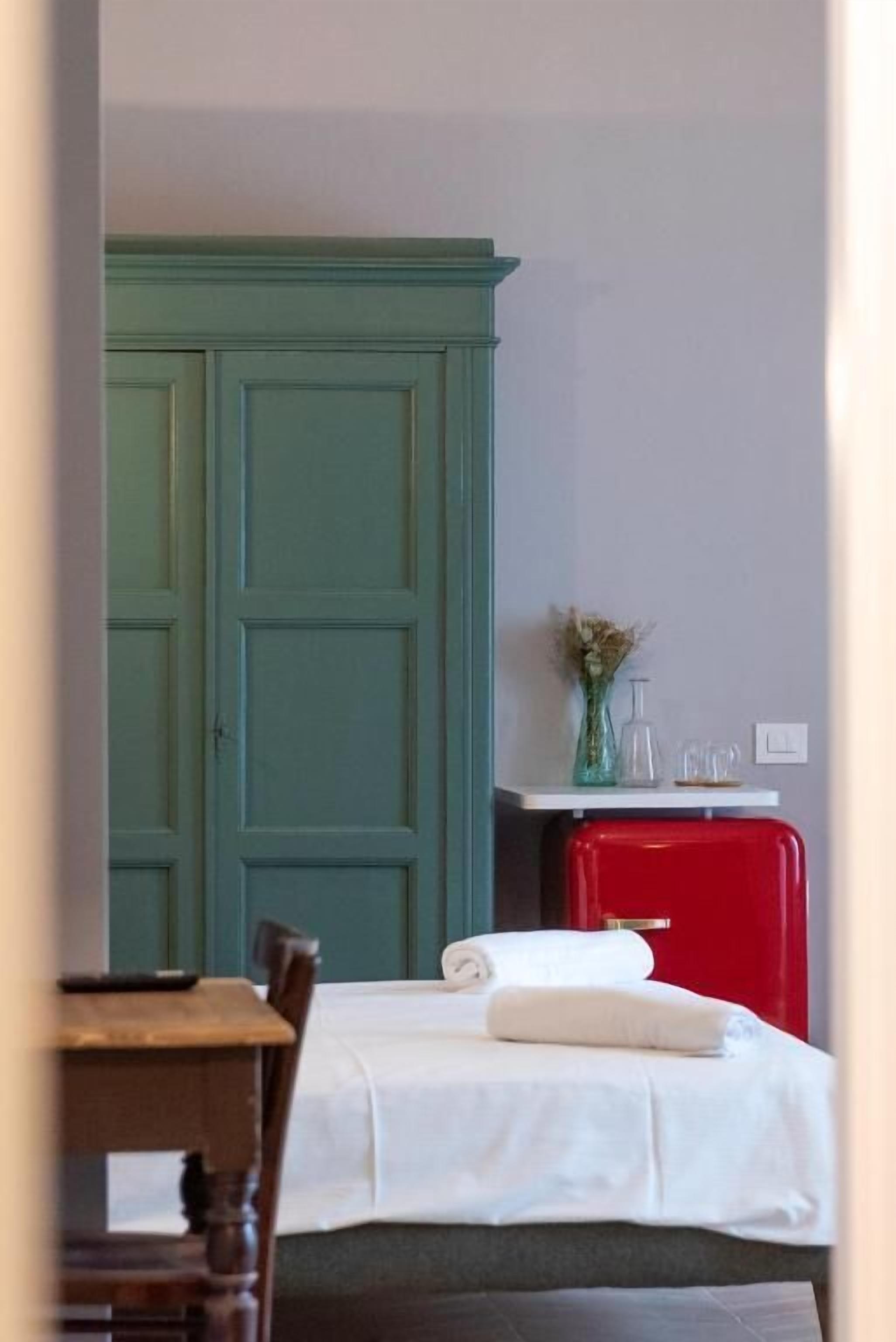 Quarto de hotel com uma cama, um aparador vermelho, um guarda-roupa verde e uma pequena mesa com um vaso de flores.
