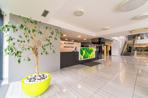 Um hall de hotel com balcão de recepção, uma grande planta em vaso e uma escada ao fundo.