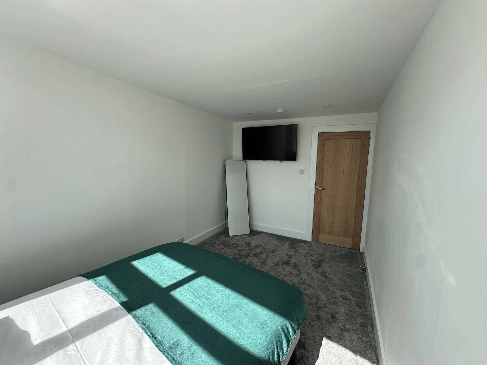 Um quarto pequeno e estreito, com uma cama, televisão fixada na parede e uma porta.