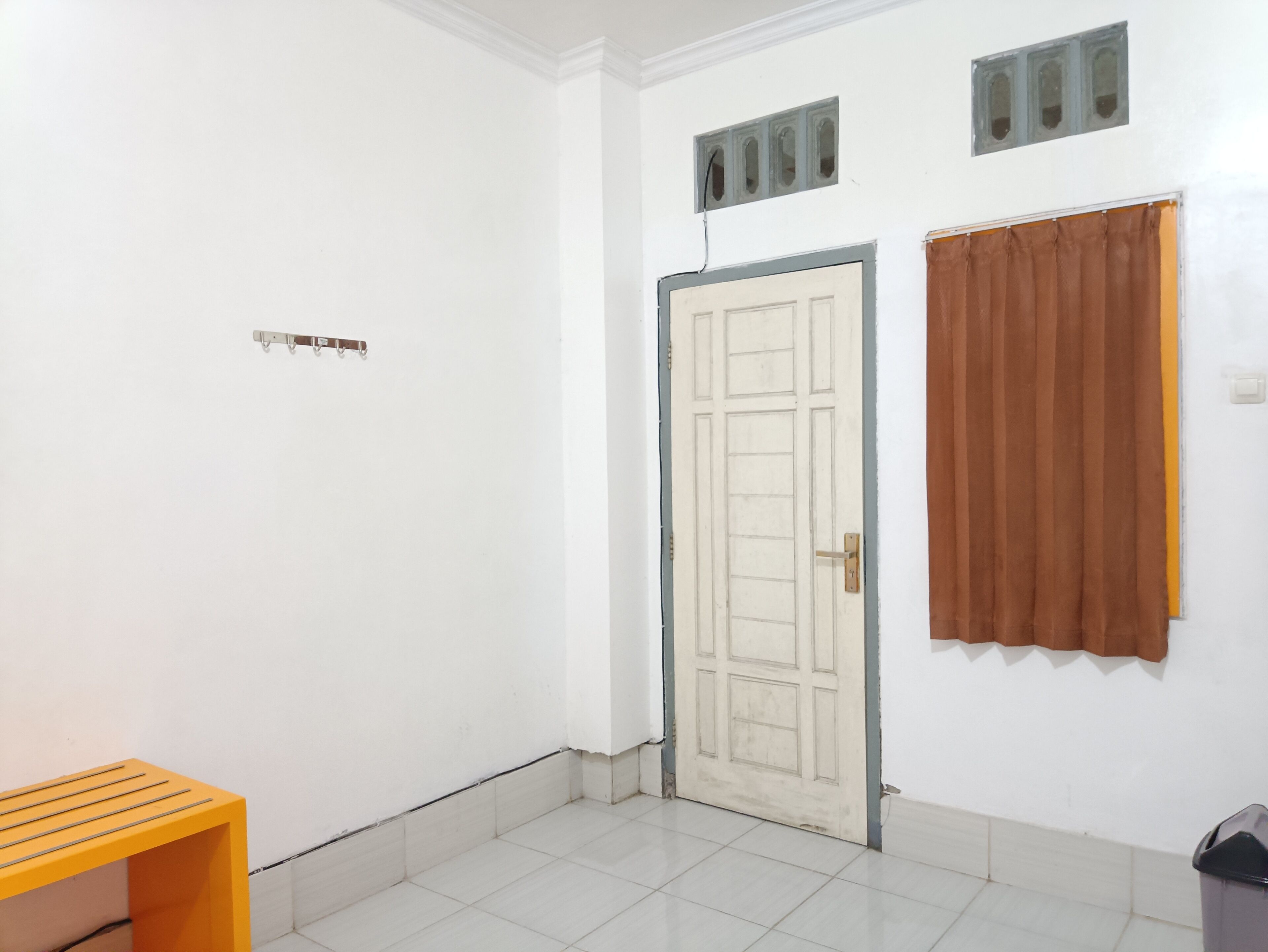 Um quarto com uma porta branca, uma pequena janela com cortina e um banco amarelo.