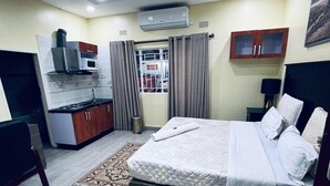 Quarto de hotel compacto com uma cama, pequeno espaço de cozinha, televisão e janela com cortinas.